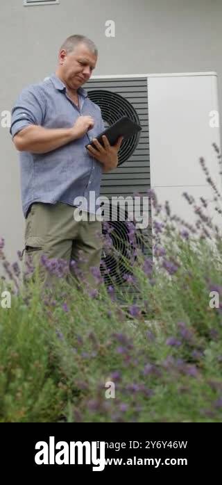 A man closely examines his external heat pump unit, consulting a tablet ...