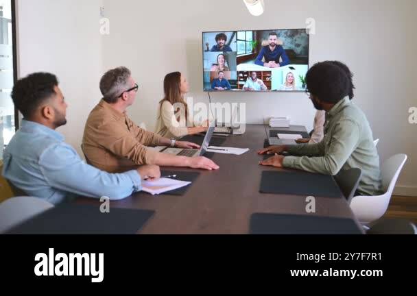 Team of professionals sits around conference table, engaging in an online video conference with ...