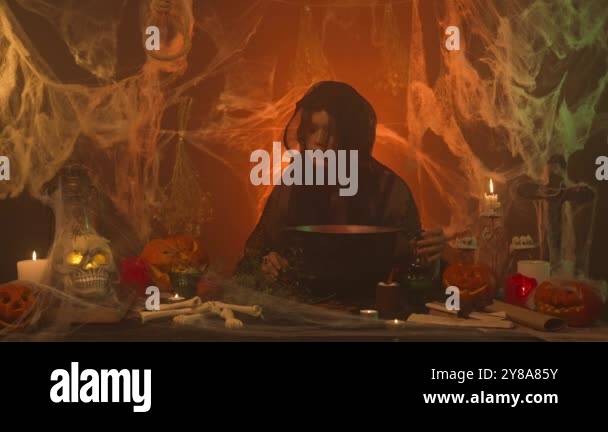 Witch in black clothing brews potion in a scary kitchen with pumpkins ...