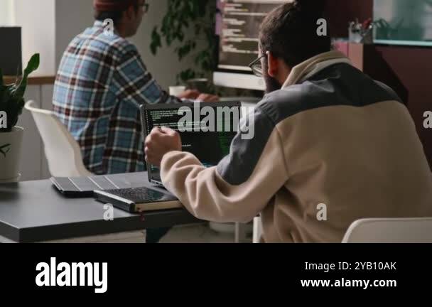 Medium rear shot of two male programmers in casual clothes sitting at desks in IT startup office ...