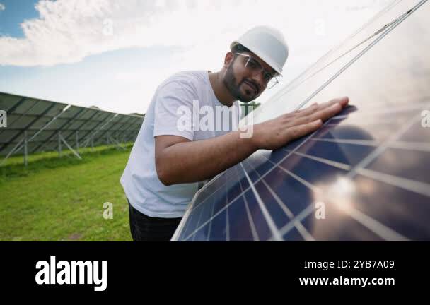 Indian engineer in a white helmet inspects solar panels closely by running his hand across the ...
