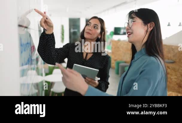 Two multiethnic women teammates stand near whiteboard review written ...