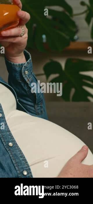 A Pregnant Woman Sits On A Sofa And Drinks Orange Juice She Is Wearing A Denim Shirt Over A