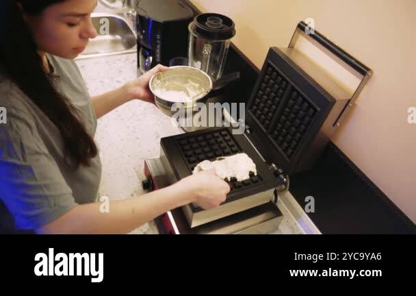 With a smile on her face, a young woman fills an electric waffle maker ...