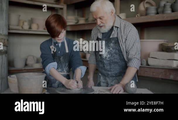 Skilled boy working with throwing wheel making pot while grandfather ...