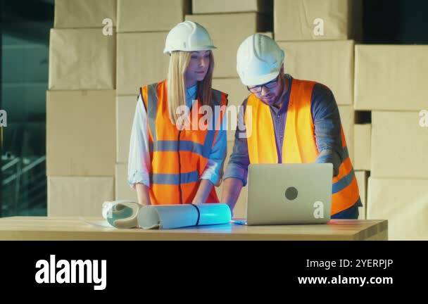Engineers in Safety Helmets and High-Visibility Vests Review Blueprints and Work on a Laptop in ...