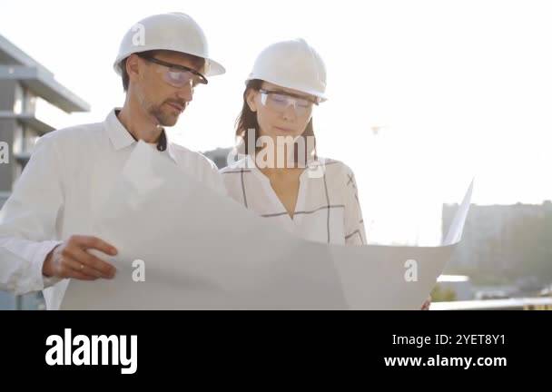 Two architects, wearing safety helmets and glasses, are examining ...