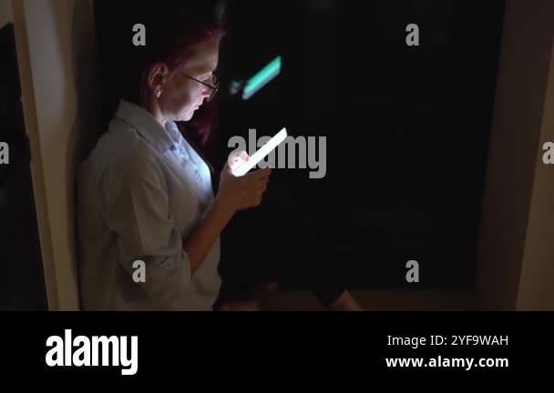 In a dim room, a young woman sits by a window, engrossed in her ...
