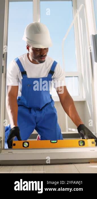Skilled technician in blue overalls and hard hat adjusts window frame ...