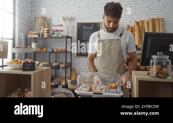 Young bearded man in an apron working in a bakery shop, arranging ...