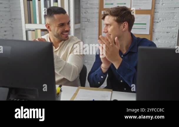 Two men clapping and smiling at their desks in a modern office setting ...