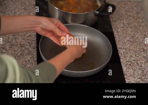A young woman breaks eggs in a frying pan on the stove preparing scrambled eggs in the kitchen ...