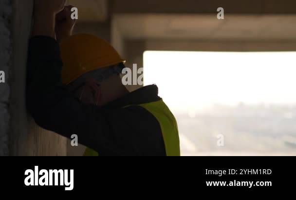 Workman knocks his hands and head against the wall. Stressed worker in hard hat stands on ...