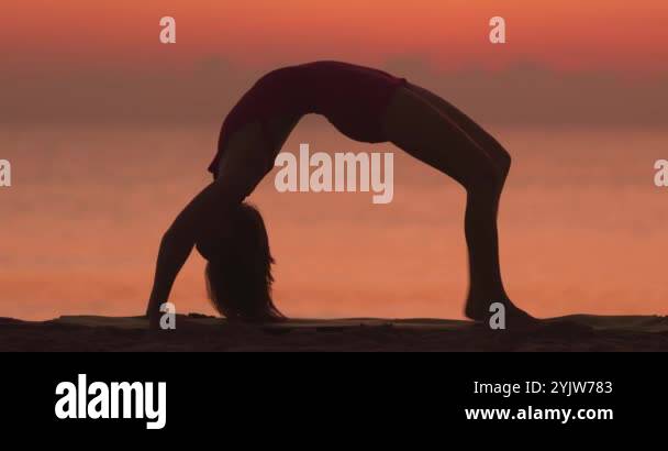 A woman performs the one-legged bridge pose in silhouette on a sandy beach at sunset, with the ...