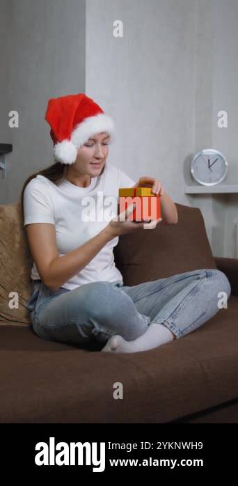 Woman in Santa hat sitting on sofa opening christmas gift. Frustration ...