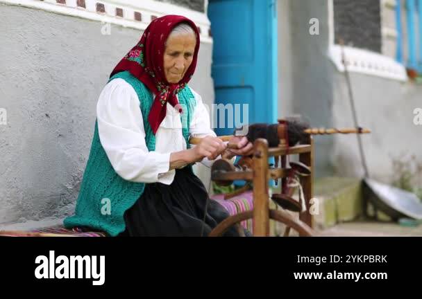 Elderly woman sits on bench and works with distaff with spinning wheel ...