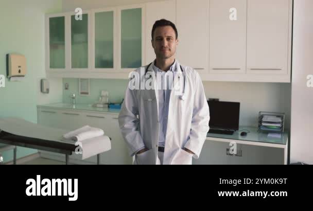 Smiling male general practitioner in white coat posing at workplace ...