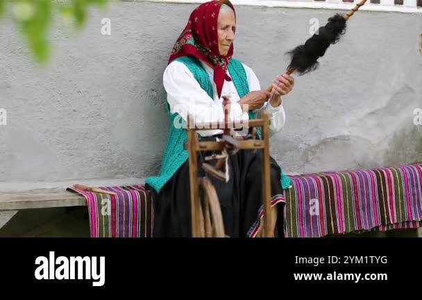 Elderly woman sits on bench and works with distaff with spinning wheel ...