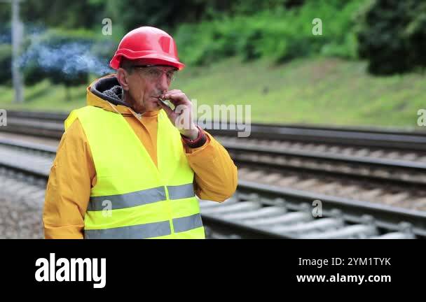 Railway man in red hard hat stands on railway track and smokes. Working ...