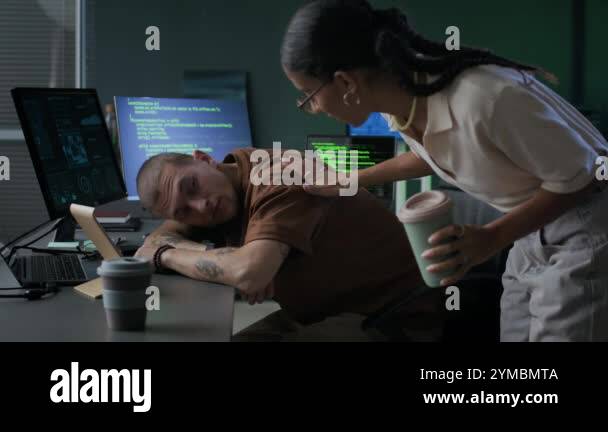 Young female biracial programmer waking up her exhausted young male Caucasian colleague sleeping ...