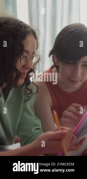 Vertical shot of two teenage girls, leaning on comfy bed, engaging in ...