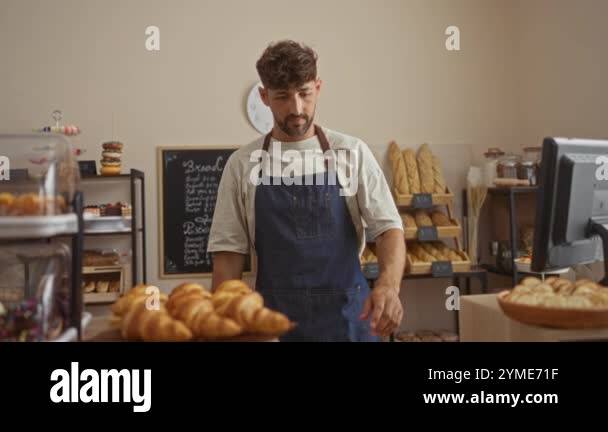 Young man in a bakery with pastries and croissants, wearing apron ...
