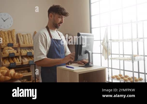 Young man in a bakery shop standing at the counter using a computer ...