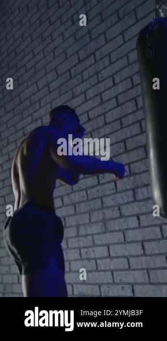 A man vigorously trains with a punching bag in a dimly lit gym ...