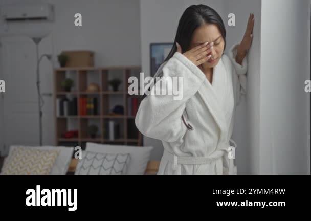 Woman feeling dizzy indoors wearing a white robe, leaning against a ...