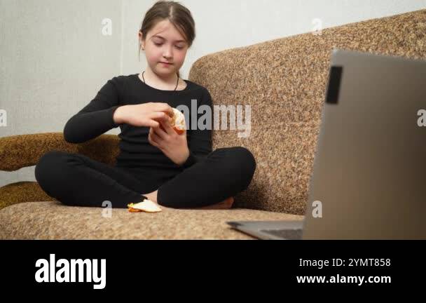Girl peeling orange while using laptop for online learning. Young student engages in virtual ...