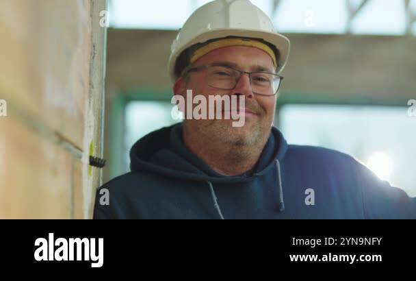 Construction worker in hard hat smiling slightly, leaning against wall ...