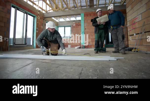 Construction worker measuring PVC pipes on floor, with two colleagues ...