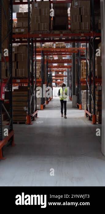 Warehouse worker wearing a safety vest and hardhat walks through ...