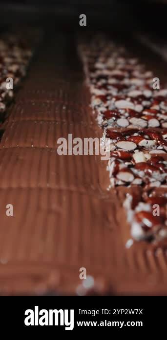 A nut bar moves along a conveyor belt and is coated with chocolate in a ...