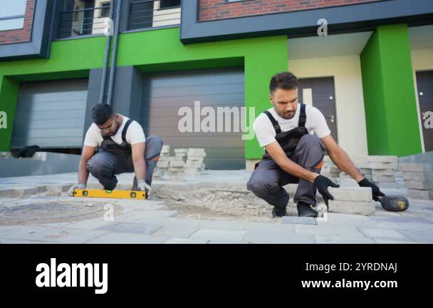 Two multiracial workers laying paving stones with precision. One uses a ...