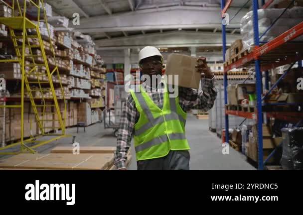 African American warehouse worker in a safety vest and hard hat walking ...