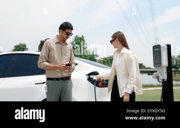 Slow motion couple using smartphone to pay EV car battery at charging station connected to power ...