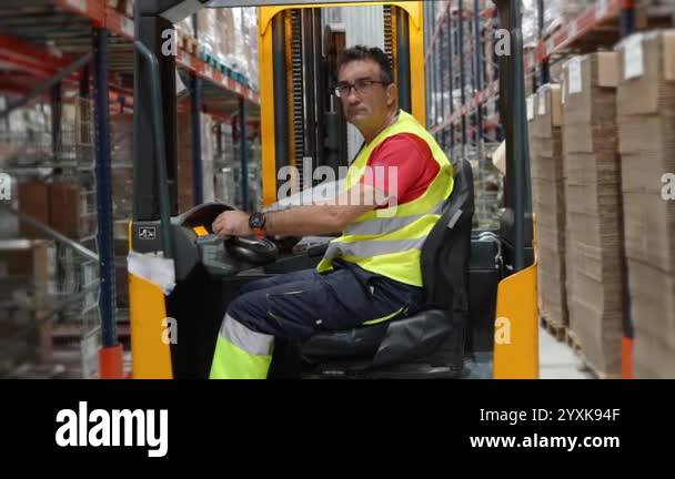 Warehouse worker wearing a safety vest is driving a forklift through a ...