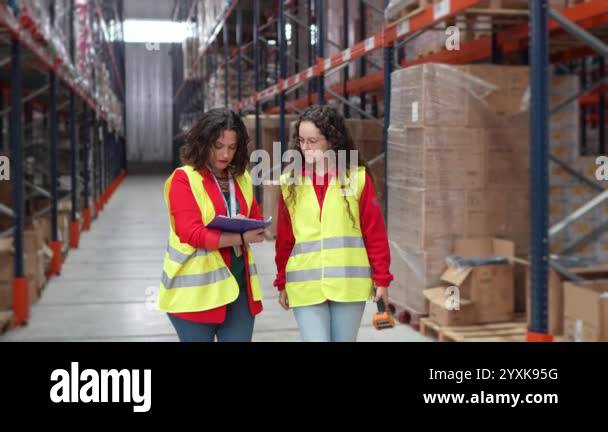 Two female warehouse workers discuss logistics while walking in slow ...