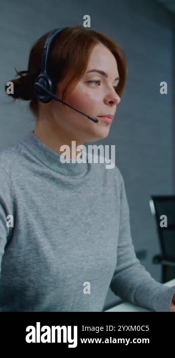 Vertical shot Caucasian female hotline operator talks on headset with customer, works on ...