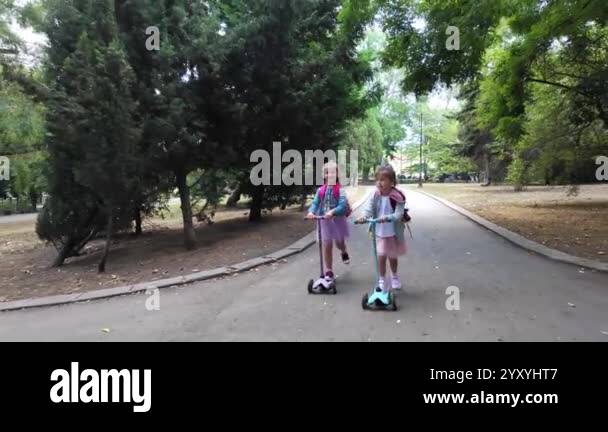 Two girls with backpacks, riding scooter in the park. Kids having fun outdoors with scooters ...