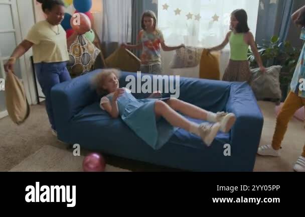 Girl falling down on sofa, four friends throwing cushions at her while ...