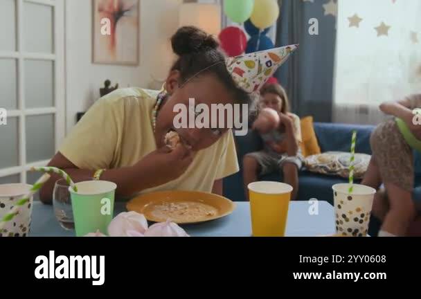 Gen alpha girl with party hat enjoying delicious cake at festive table while friends playing in ...