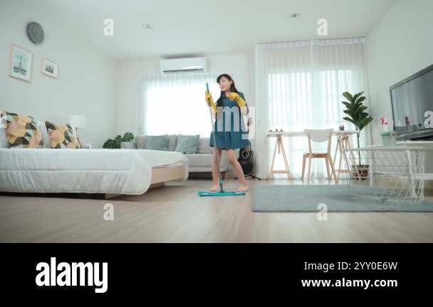 Attractive cute daughter mopping floor while moving to music at modern house. Energetic girl ...