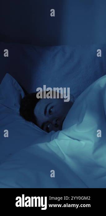 Vertical video. Man lies under blanket in bed looking concerned ...