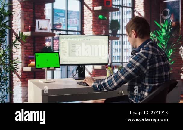 Man In Home Office Looks At Green Screen Tablet And Computer Typing And Executing Programming