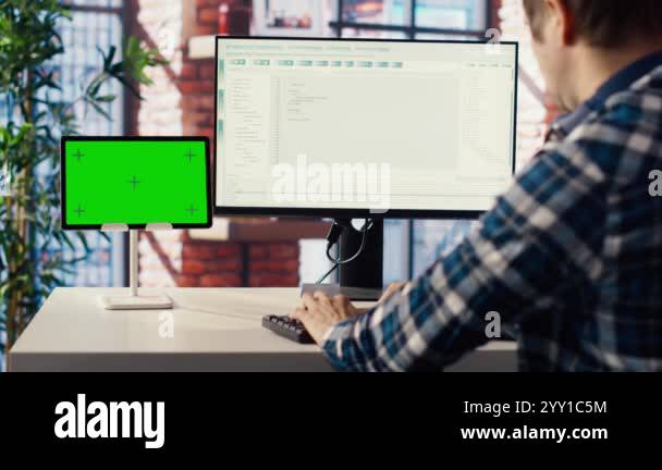 Man In Home Office Looks At Green Screen Tablet And Computer Typing And Executing Programming