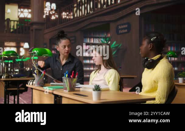 Caucasian tutor guiding elementary school girls at a library desk ...