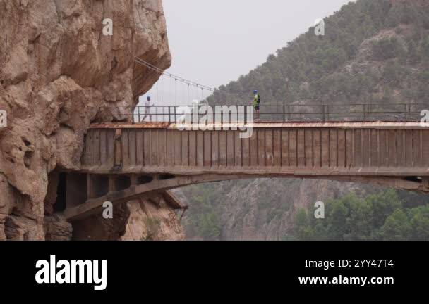 Two individuals carefully traverse a rugged bridge spanning a deep ...