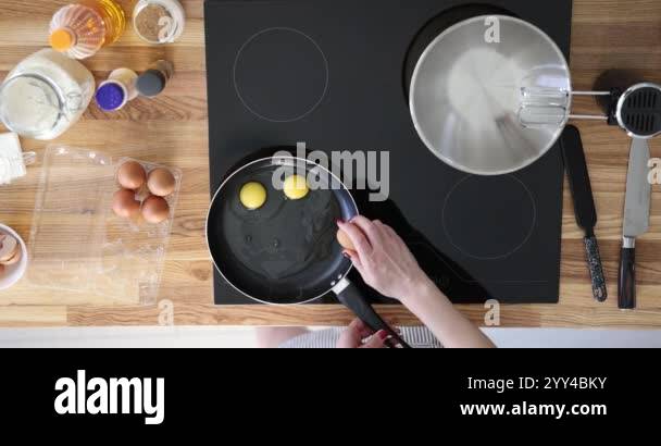 Woman breaking eggs in a frying pan on an induction cooker top view. Housewife preparing food ...